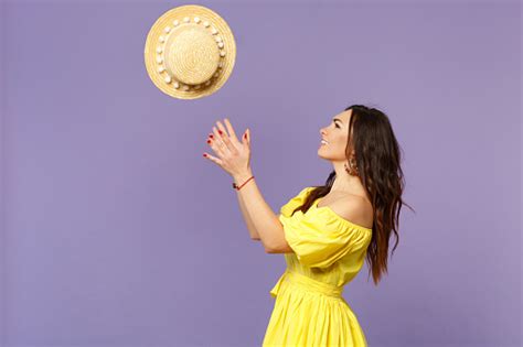 스튜디오에서 파스텔 바이올렛 벽 배경에 고립 된 여름 모자를 던지는 노란 드레스에 웃는 매력적인 젊은 여자의 측면 보기 사람들은 진심으로 감정 라이프 스타일 개념입니다 복사