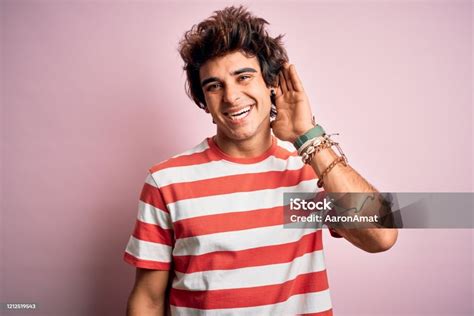 줄무늬 캐주얼 티셔츠를 입은 젊은 잘생긴 남자는 소문이나 험담에 귀를 듣고 귀에 손으로 미소 고립 된 분홍색 배경 위에 서 청각 장애 개념입니다 귀 부분에 대한 스톡 사진
