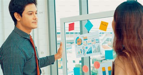 비즈니스 사람들은 사무실의 프로젝트 계획 보드에서 일하고 동료 친구와 대화하여 프로젝트 개발을 분석합니다 그들은 유리 벽에 게시 된 스티커 메모를 사용하여 정리합니다