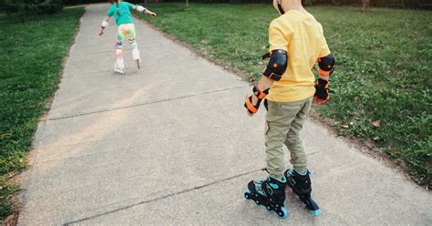 헬멧을 쓴 친구 소년과 소녀는 여름날 공원에서 롤러 스케이트를 타고 있습니다 자매는 형제가 롤러 스케이트를 타도록 자극하도록 격려합니다 형제 자매 경쟁 계절에 따라