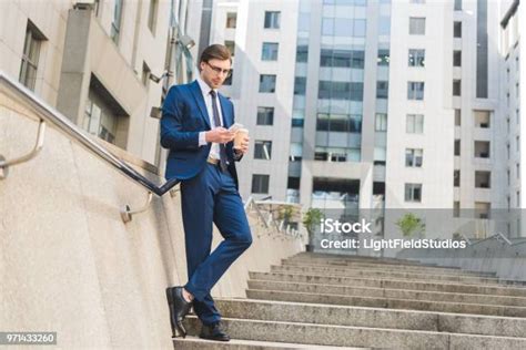 종이 컵 커피 비즈니스 빌딩 근처 계단에 스마트폰을 사용 하 여 세련 된 정장에 매력적인 젊은 사업가 수트에 대한 스톡 사진 및