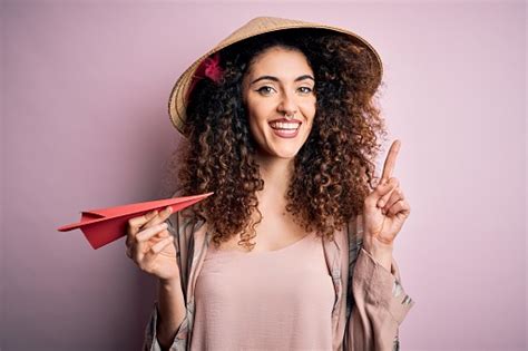 젊은 아름 다운 여자 와 곱슬 머리 와 피어싱 착용 아시아 모자 들고 종이 비행기 와 함께 놀란 아이디어 또는 질문 가리키는 손가락 으로 행복한 얼굴 번호 하나 가리키기에