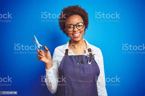 행복한 얼굴 서서 치아를 보여주는 자신감 미소와 함께 미소와 파란색 고립 된 배경 위에 가위를 들고 아프리카 계 미국인 미용사 여자 헤어 스타일리스트에 대한 스톡 사진 및