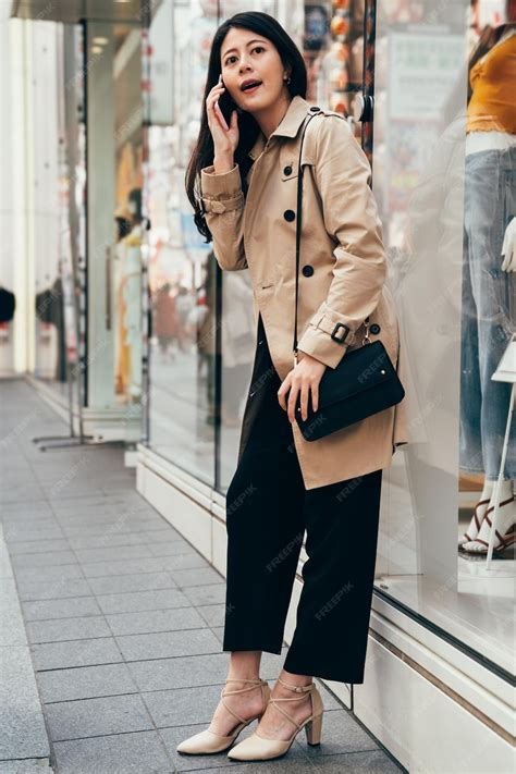 휴대폰으로 이야기 하는 친구를 기다리는 사무실 여자의 전체 길이 젊은 여성 사업가가 일본 도톤보리에 있는 옷가게에서 쇼핑 창가 근처에 서서 일을 마치고 만납니다