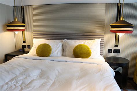 노란색 벨루어 겹겹이 쌓인 흰색 베개에 기대어 있는 원형 쿠션 질감이 있는 더블 침대 배턴 헤드보드 접힌 이불 스탠드의 디지털 알람 시계 조명이 켜진 교수형 펜던트 램프