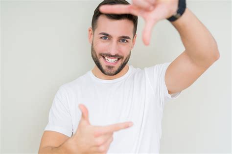 손으로 만드는 프레임 및 행복 한 얼굴로 손가락을 웃 고 고립 된 배경 위에 캐주얼 티셔츠를 입고 젊은 잘생긴 남자 창의력과