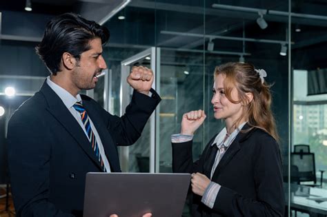 사무실에 서서 노트북을 들고 미소를 지으며 주먹을 치켜들고 팀 성취를 축하하는 사업가와 사업가 성공 동기 부여 행복 및 자신감 있는 긍정적인 팀워크 제스처를 함께 보여줍니다
