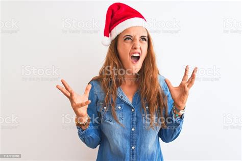 고립 된 배경에 크리스마스 모자를 쓰고 있는 젊은 아름다운 빨간 머리 여자는 미친 미친 듯이 외치고 공격적인 표정과 팔을 들고 고함을 쳤습니다 좌절 개념입니다 감정에 대한