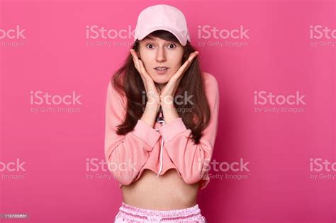 넓은 열린 눈을 가진 예쁜 갈색 머리 여성 로 지 짧은 까마귀와 모자를 착용 뺨에 손바닥을 유지 모델은 고립 된 분홍색 배경에 서 서 카메라를 보고 스튜디오에서 놀란 십 대