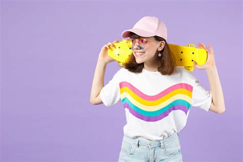 비 비드 한 옷에 웃는 십 대 소녀 노란 스케이트 보드를 들고 심장 안경 보라색 파스텔 벽 배경에 고립 된 찾고 사람들은 진심으로 감정 라이프 스타일 개념입니다 복사 공간을