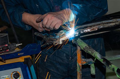 클로즈업 보기 강한 남자는 팔에 장갑 없이 파란색 작업 바지에 용접기 금속 제품은 차고 워크샵에서 용접 기계로 용접 블루 연기와 오렌지 스파크 멀리 날아 강철에 대한 스톡