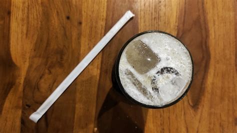 우유를 넣은 아이스 커피 한 잔이 빨대와 함께 나무 테이블 위에 놓여 있습니다 음료는 상단에 거품이 많고 얼음 조각이 보여