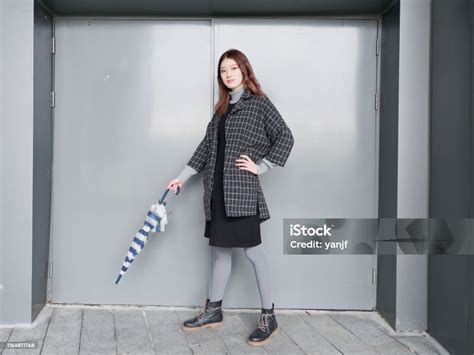 회색 철문 앞에서 포즈를 취하고 허리에 손을 얹고 우산을 걷는 막대기로 들고 있는 아름다운 긴 머리를 가진 젊은 아시아 여성의 초상화 20 29세에 대한 스톡 사진 및 기타