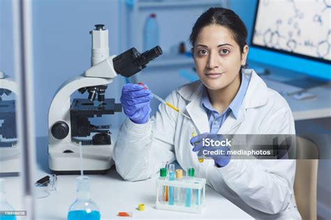 실험실 코트와 수술 장갑에 젊은 인도 여성 약사의 초상화 책상에 앉아 파이펫을 사용 하 여 테스트 튜브에 시약을 혼합 Stem 주제에 대한 스톡 사진 및 기타 이미지