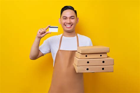 갈색 앞치마를 입은 젊은 아시아 남자 바리스타 바텐더 직원이 노란색 배경에 골판지와 신용 카드 판지에 이탈리아 피자를 들고