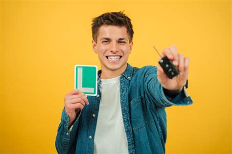 활기찬 노란색 배경에 자동차 열쇠와 새로운 운전자 L 표지판을 들고 흥분한 젊은이 백인 남자는 모자를 축하하고 그는 색상 배경에서 운전 면허증을 취득했습니다 자동 학습자