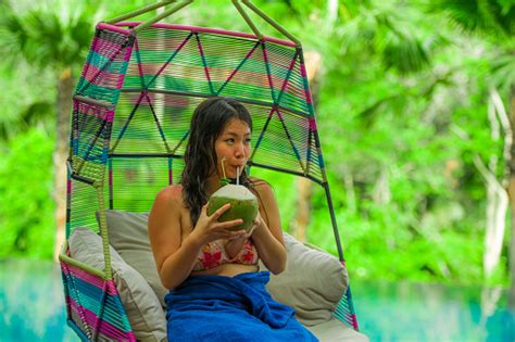 젊은 행복 하 고 매력적인 아시아 중국 여자 걸려 열 대 섬에 럭셔리 휴가 즐기는 리조트 수영장에서 편안한 코코넛 물 마시는 해먹에 앉아 고급 호텔에 대한 스톡 사진 및