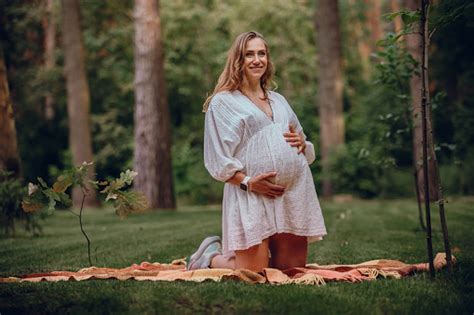 하얀 보헤미안 여름 드레스를 입은 임산부는 숲 배경 근처에서 임신한 배에 손을 얹고 담요 위에 앉아 화창한 날에 귀여운 미소를 짓고 있다 라이프스타일 임신 개념 여유 공간