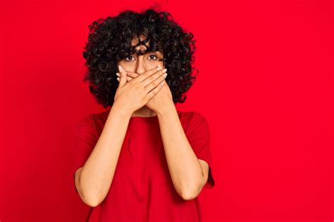 고립 된 빨간 배경 위에 캐주얼 티셔츠를 입고 곱슬 머리를 가진 젊은 아랍 여성은 실수로 손으로 입을 덮는 충격을 받았습니다 비밀 개념입니다 T 셔츠에 대한 스톡 사진 및