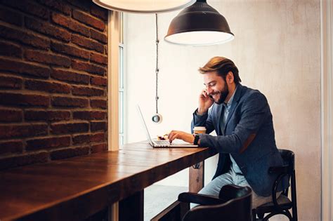도시 카페에서 노트북을 가지고 일하는 젊은 미소 짓는 사업가 커피를 마시고 전화로 이야기 양복과 수염을 착용 가방에 대한 스톡