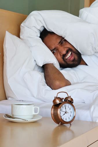 베개를 덮고 귀를 덮고 더블 침대에 누워 있는 심술궂은 인도 남자의 이미지 침대 옆 탁자에 알람 시계 청동 금속 케이스 복고풍 알람 시계 더블 벨 아날로그 디스플레이 시계