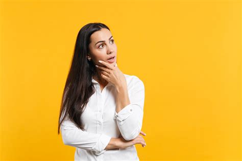 흰색 셔츠에 잠겨있는 매력적인 젊은 여자의 초상화 턱에 손을 넣어 스튜디오에서 노란 오렌지 벽 배경에 고립 된 옆으로 찾고 사람들 라이프 스타일 개념입니다 복사 공간을 모의