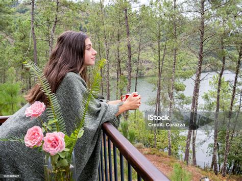 아름다운 백인 여성이 뜨거운 커피를 들고 마시고 발코니에 서서 호수와 녹색 자연을 바라 보는 초상화 이미지 마시기에 대한 스톡