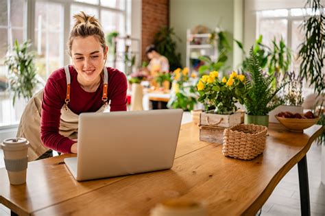 젊은 여성 플로리스트는 꽃이 만발한 꽃꽂이와 쾌활한 분위기의 활기찬 꽃집 환경에서 노트북으로 작업을 즐깁니다 2명에 대한 스톡 사진 및 기타 이미지 Istock