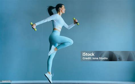 파란색 운동복을 입은 백인 젊은 여성이 피트니스 수업에서 평범한 회색 벽에 덤벨을 들고 점프를 한다 포니테일을 하고 스포츠를 하는 키 큰 갈색 머리 유럽 소녀 건강한 라이프