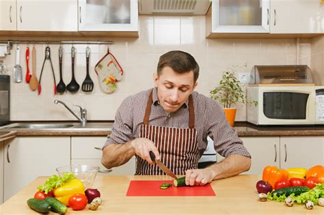 잘생긴 웃는 백인 젊은이 앞치마 테이블에 앉아 갈색 셔츠에에서 밝은 부엌에서 칼으로 샐러드 야채를 잘라냅니다 다이어트 개념입니다 건강 한 생활양식입니다 집에서 요리 음식