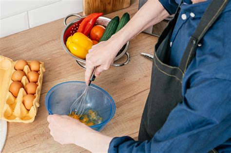 파란색 긴소매 셔츠를 입은 인식할 수 없는 남자의 손이 파란색 플라스틱 그릇에 달걀을 때려 놓고 테이블에 와이어 거품기로 말했습니다 채소에 대한 스톡 사진 및 기타 이미지