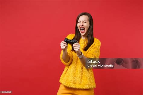 노란 모피 스웨터에 미친 젊은 여자의 초상화 비명 밝은 빨간색 벽 배경에 고립 된 조이스틱으로 비디오 게임을 재생 사람들은