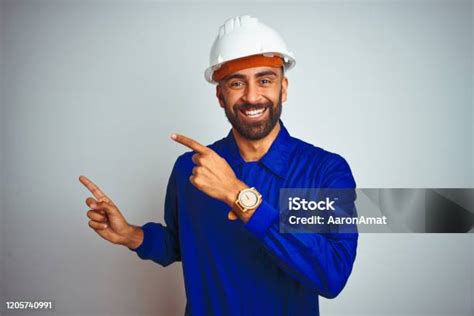 잘 생긴 인도 노동자 남자 는 고립 된 흰색 배경 위에 유니폼과 헬멧을 입고 미소와 측면에 두 손과 손가락으로 가리키는 카메라를 보고 가리키기에 대한 스톡 사진 및 기타