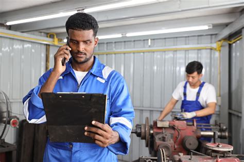 금속 선반 기계를 사용하는 다민족 기술자 정비사는 차고에서 스마트 휴대폰과 클립 보드로 자동차 디스크 브레이크 연마를 작동합니다