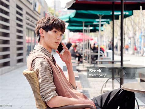 격자 무늬 셔츠와 울 조끼에 곱슬 검은 머리를 가진 잘 생긴 중국 청년의 초상화는 화창한 날에 손에 자신의 휴대 전화와 야외 카페앉아 남성 패션 시원한 아시아 젊은 남자