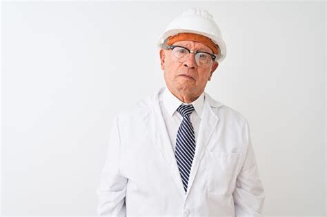 얼굴에 심각한 표정으로 고립 된 흰색 배경 위에 코트와 헬멧을 입고 선임 회색 머리 엔지니어 남자 카메라를 보는 간단하고 자연스러운 건축가에 대한 스톡 사진 및 기타 이미지