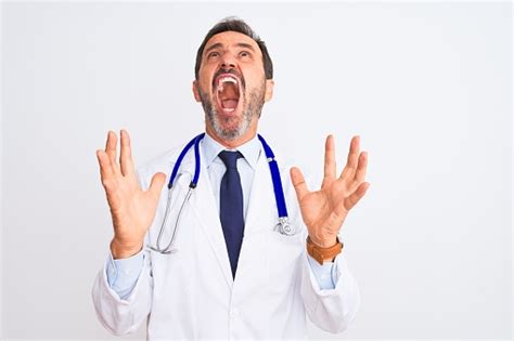 외투와 청진기를 입은 중년의 의사 남자가 고립 된 흰색 배경 위에 서서 미친 듯이 외치고 공격적인 표정과 팔을 들고 소리쳤다 좌절 개념입니다 감정에 대한 스톡 사진 및 기타