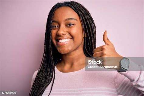 고립 된 배경 위에 분홍색 스웨터를 입고 행복한 엄지 손가락으로 손을 들고 제스처를 하는 젊은 아프리카 계 미국인 여성의 클로즈업 성공을 보여주는 카메라를 보고 표정을