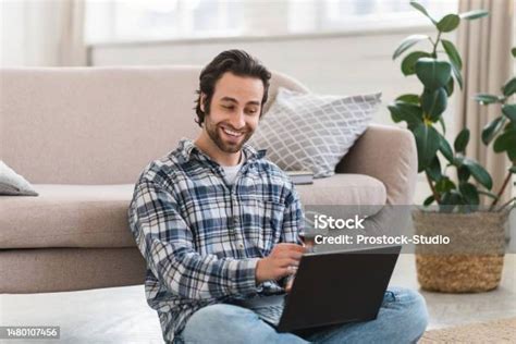 수염이 있는 국산 옷을 입은 쾌활한 밀레니얼 유럽 남자가 바닥에 앉아 컴퓨터와 함께 와인 한 잔으로 환호한다 20 24세에 대한 스톡 사진 및 기타 이미지 Istock