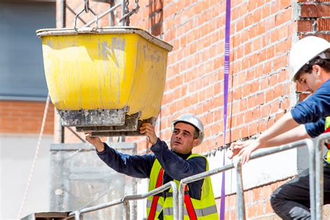 보호 및 안전 장비를 착용한 작업자가 높이에서 크레인 체인에 콘크리트 혼합물이 있는 컨테이너를 들어 올리도록 지시합니다 2명에