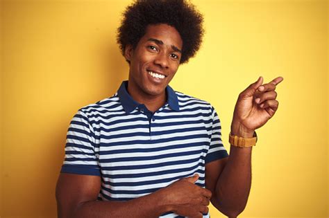 해군 줄무늬 폴로를 입은 미국 남자는 얼굴에 큰 미소를 지으며 고립 된 노란색 배경 위에 서서 카메라를 보고 있는 옆구리를 손가락으로 가리킵니다 가리키기에 대한 스톡 사진