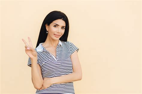 매력적인 갈색 머리 여성의 스튜디오 샷 기쁘게 표현 제스처 실내 및 평화 기호를 보여줍니다 메이크업과 깨끗한 순수한 피부를 가지고 캐주얼 옷을 입고 스튜디오 벽에 고립 25