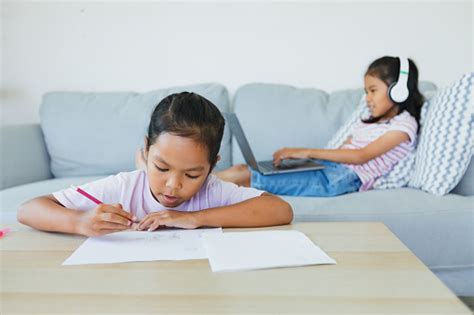 아시아 의 어린이 소녀는 소파에 앉아 교사와 수업 그룹과 화상 회의 채팅을하는 그녀의 여동생과 워크 시트를하고 아이들은