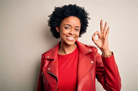 젊은 아름 다운 아프리카 계 미국인 Afro 여자 곱슬 머리 캐주얼 빨간 재킷을 입고 미소 긍정적인 하 고 확인 기호 와 손과 손가락 성공적인 식입니다 곱슬 머리에 대한