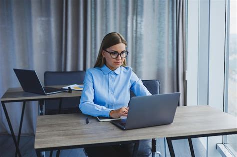 A Beautiful Woman In Glasses Sits At A Table In The Office And Works At A Laptop Corporate Work