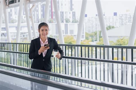 검은 양복을 입은 젊은 아시아 사업가가 전화를 사용하고 커피 한 잔을 들고 복사 공간이 있는 고가 산책로 역에 서 있습니다 30 34세에 대한 스톡 사진 및 기타 이미지