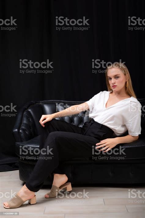 흰색 셔츠와 검은 색 패브릭 바지를 입은 젊고 매력적인 금발 모델이 배경에 검은 커튼이있는 검은 가죽 소파에 앉아 있습니다 20 24세에 대한 스톡 사진 및 기타 이미지