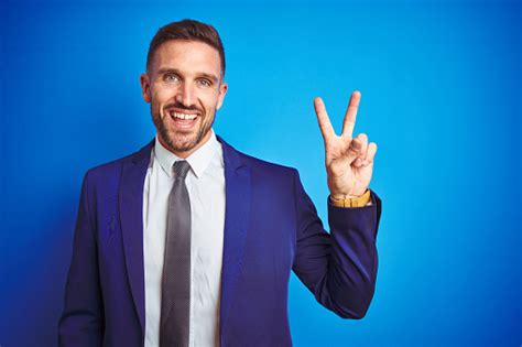 파란색 고립 된 배경을 통해 젊은 잘 생긴 사업가 는 승리 기호를하고 카메라에 윙크 행복한 얼굴미소 2번 경영자에 대한 스톡