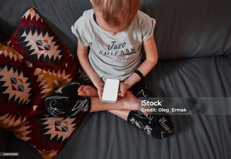 거실의 소파에 앉아있는 귀여운 아이 소녀는 스마트 폰을 사용하고 온라인 게임을합니다 전화를 들고 화면 장치를보고 아이는 소파에 앉아서 거실에서 휴식을 취합니다 기술 개념