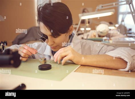 Concentrated Young Female Watchmaker In Lab Coat Sitting At Table And Using Magnifying Glass